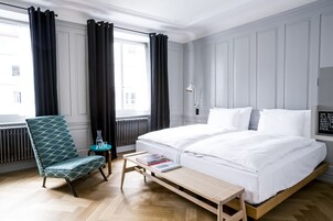 Quarto de hotel com uma cama grande, uma cadeira, uma mesinha e uma janela com cortinas.