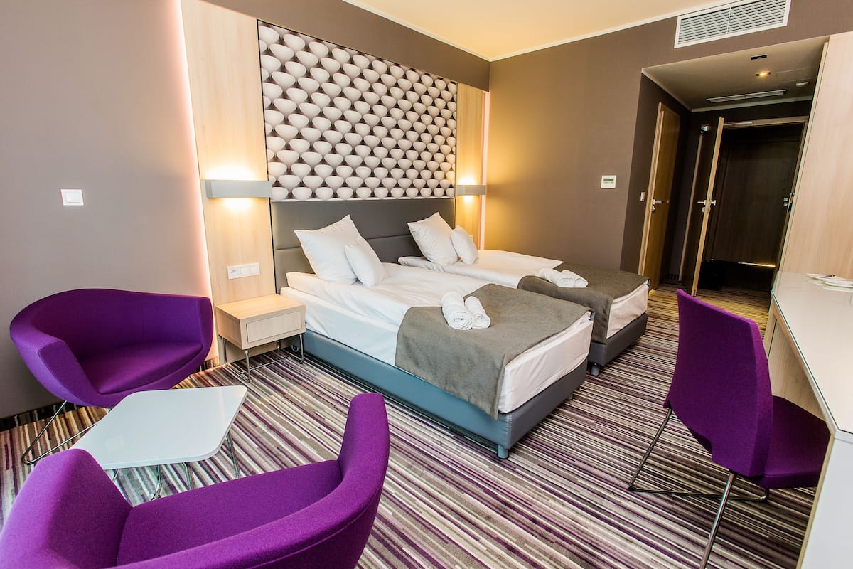 Quarto de hotel com uma cama, duas cadeiras roxas, uma mesinha, uma parede com padrão geométrico e uma porta que dá para outro cômodo.