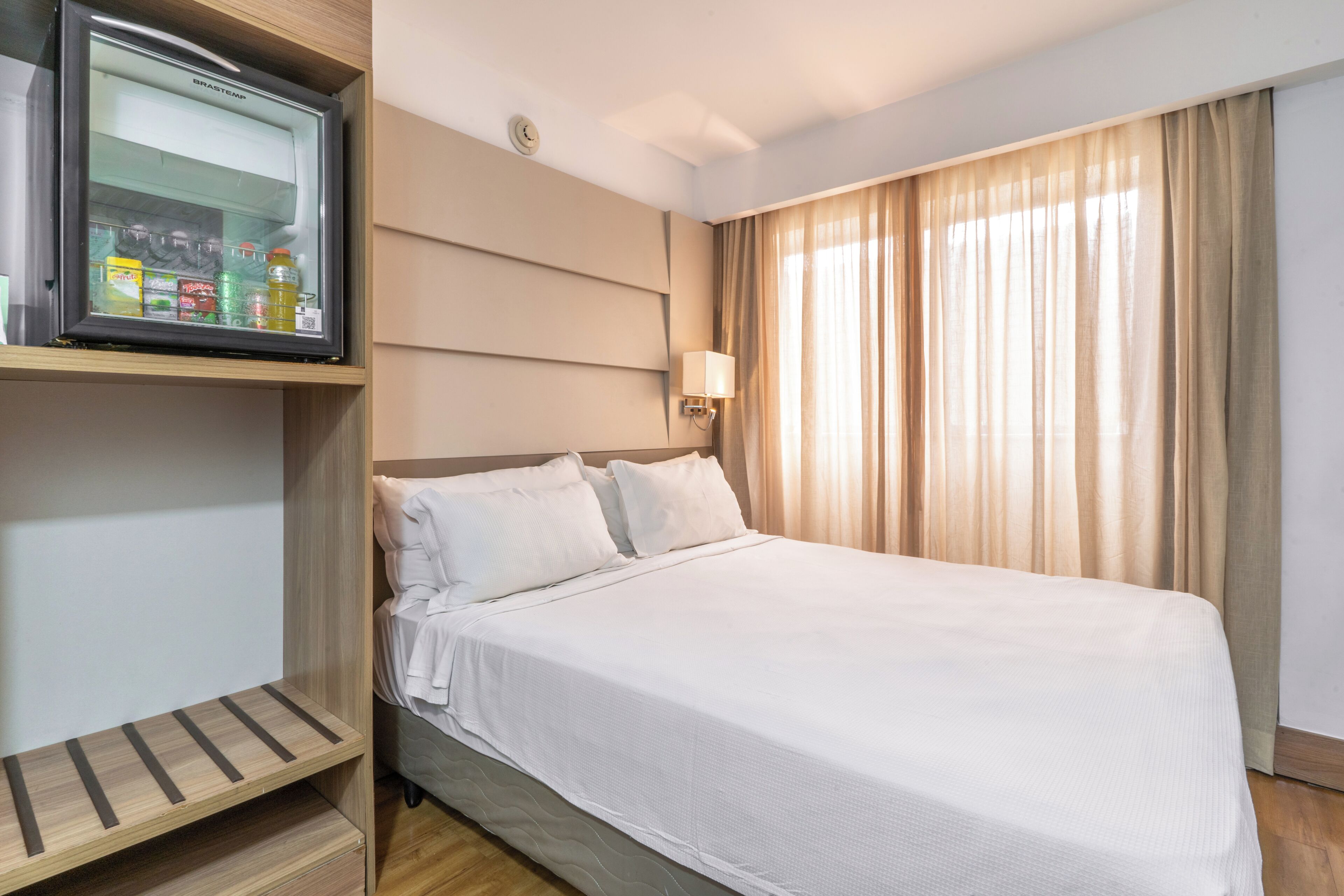Quarto de hotel com cama, televisão em prateleira, minibar e janela com cortinas.
