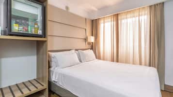 Quarto de hotel com cama, televisão em prateleira, minibar e janela com cortinas.