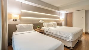 Quarto de hotel com duas camas, uma mesinha de cabeceira com abajur, uma pequena planta e uma porta de madeira.