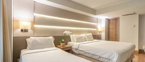Quarto de hotel com duas camas, uma mesinha de cabeceira com abajur, uma pequena planta e uma porta de madeira.
