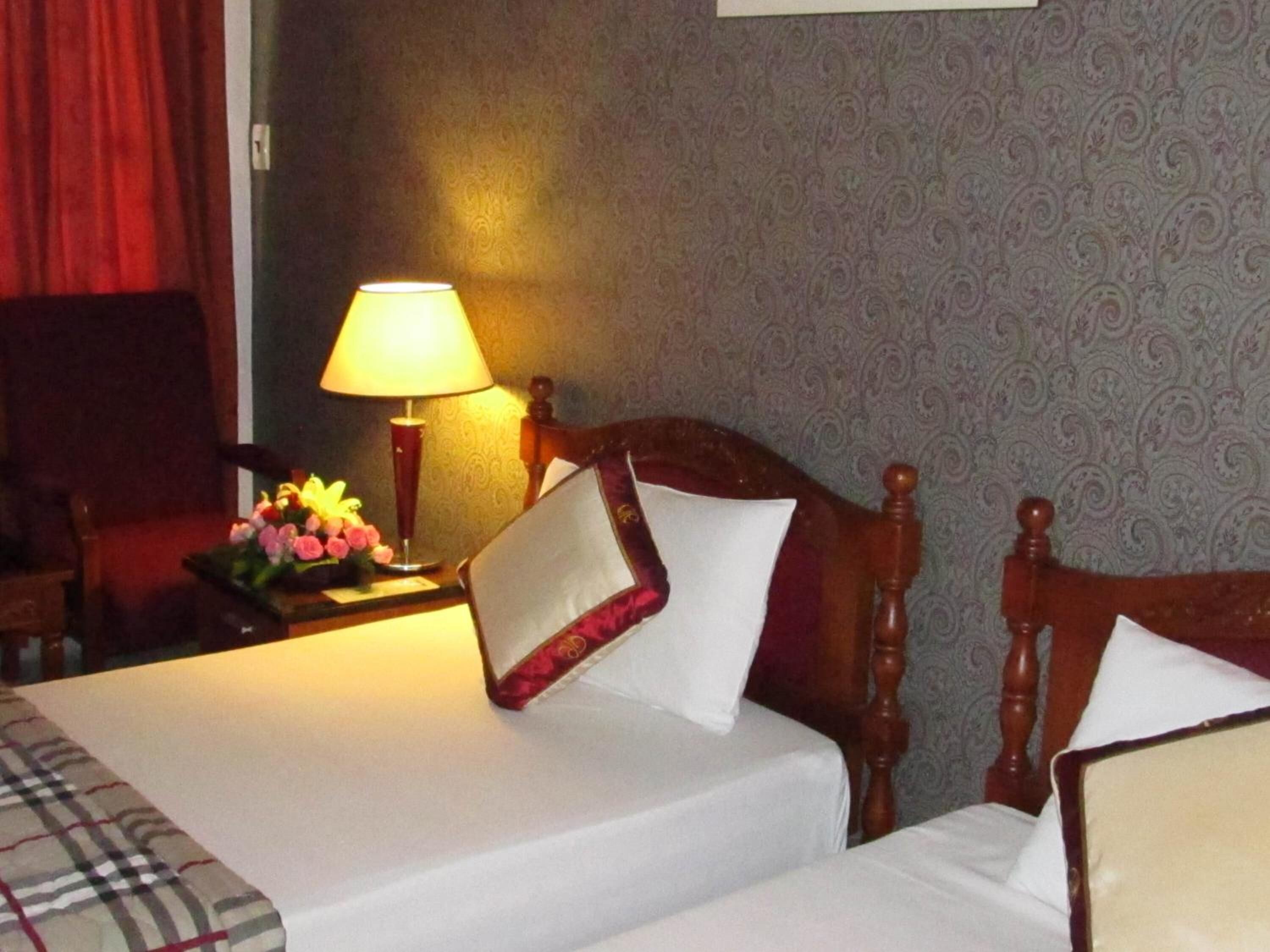 Quarto de hotel com duas camas, uma mesa de cabeceira com abajur e um arranjo floral.