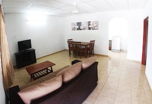 Uma sala de estar com sofá, mesa de centro, televisão, mesa de jantar com cadeiras e geladeira.