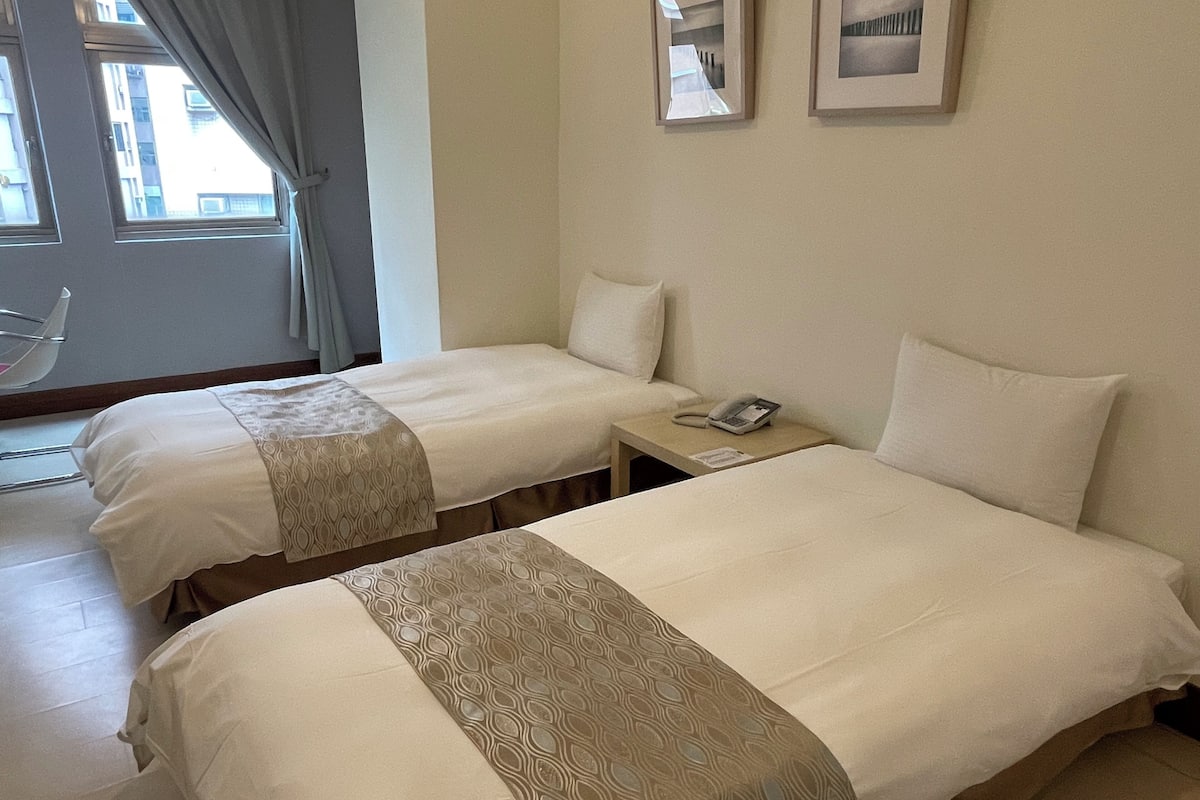 Quarto de hotel com duas camas, uma mesa de cabeceira com telefone e uma janela com cortinas.