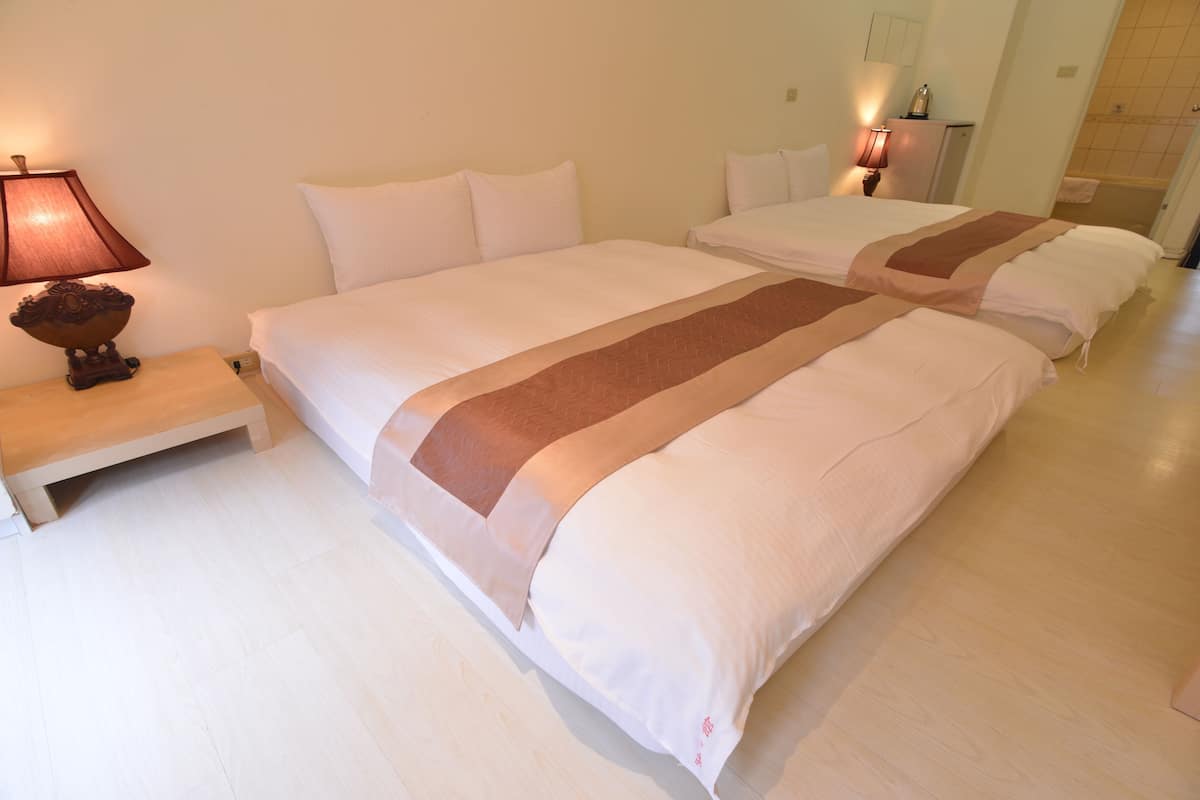 Quarto de hotel com duas camas, uma mesa de cabeceira com abajur e um banheiro visível ao fundo.