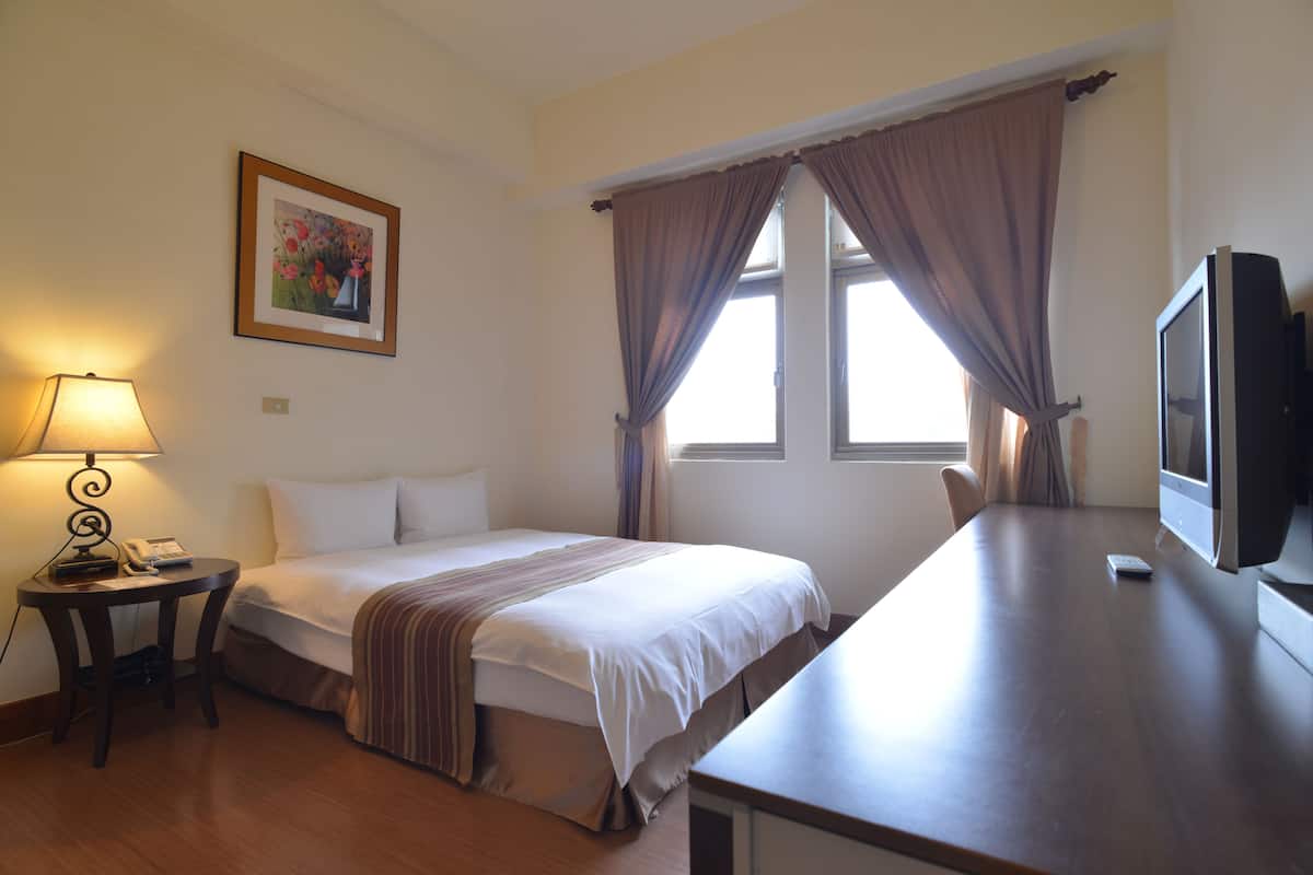 Quarto de hotel com cama, mesa de cabeceira com abajur, televisão e janela com cortinas.