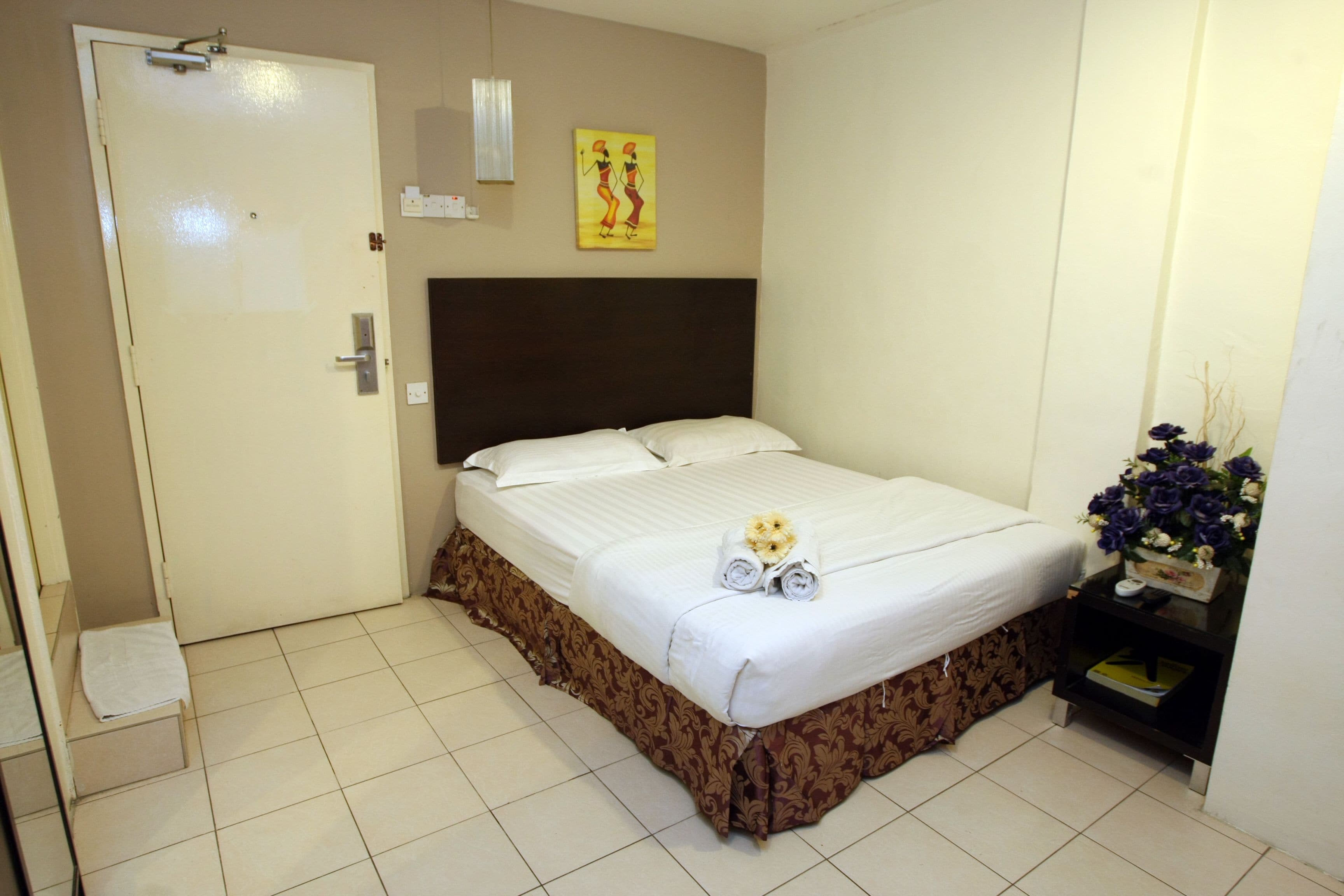 Quarto de hotel com uma cama, uma mesa de cabeceira com um vaso de flores, um quadro na parede e uma porta.