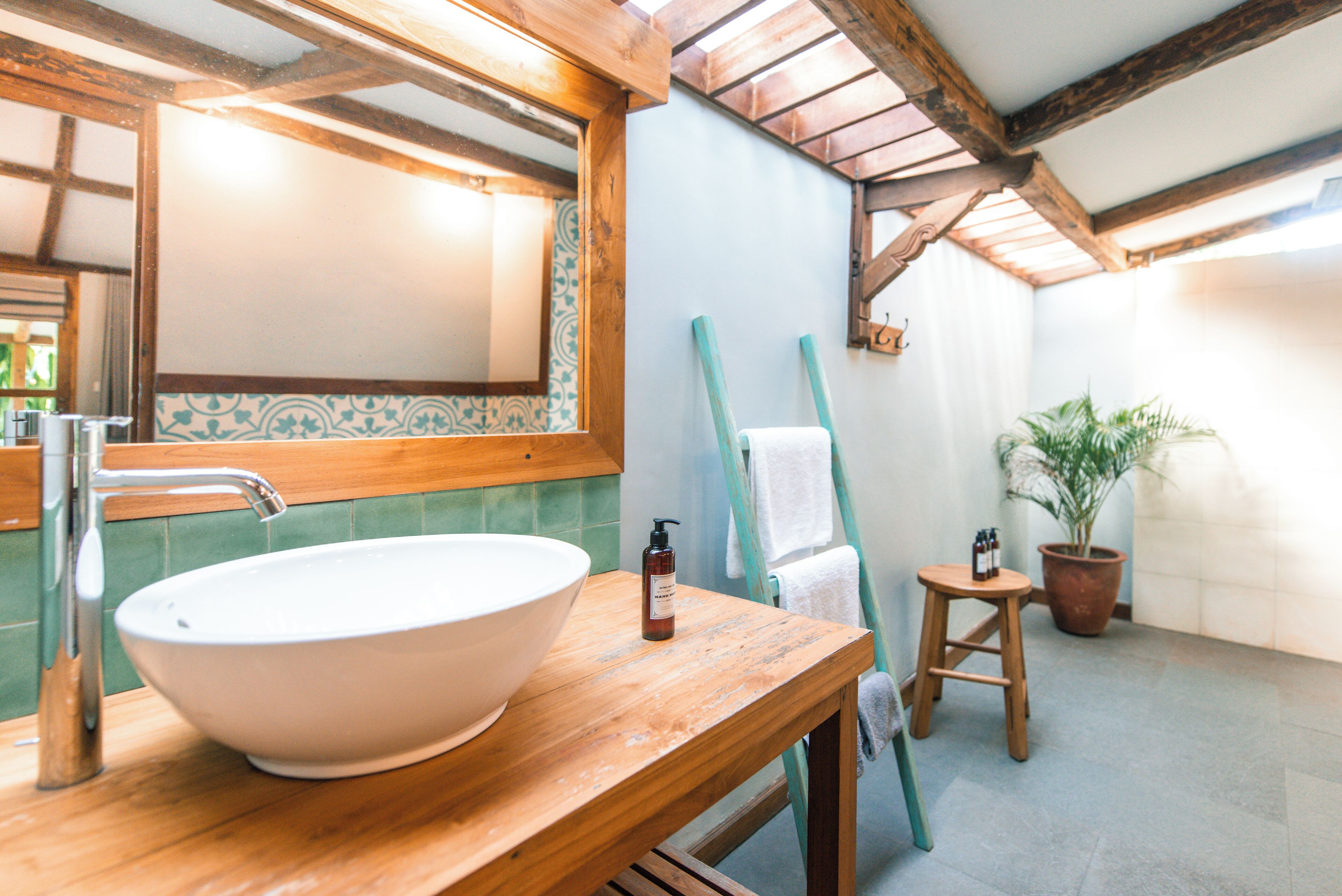 Uma casa de banho com uma grande pia branca, toucador de madeira, um espelho, um porta-toalhas, um banquinho pequeno e uma planta em vaso.
