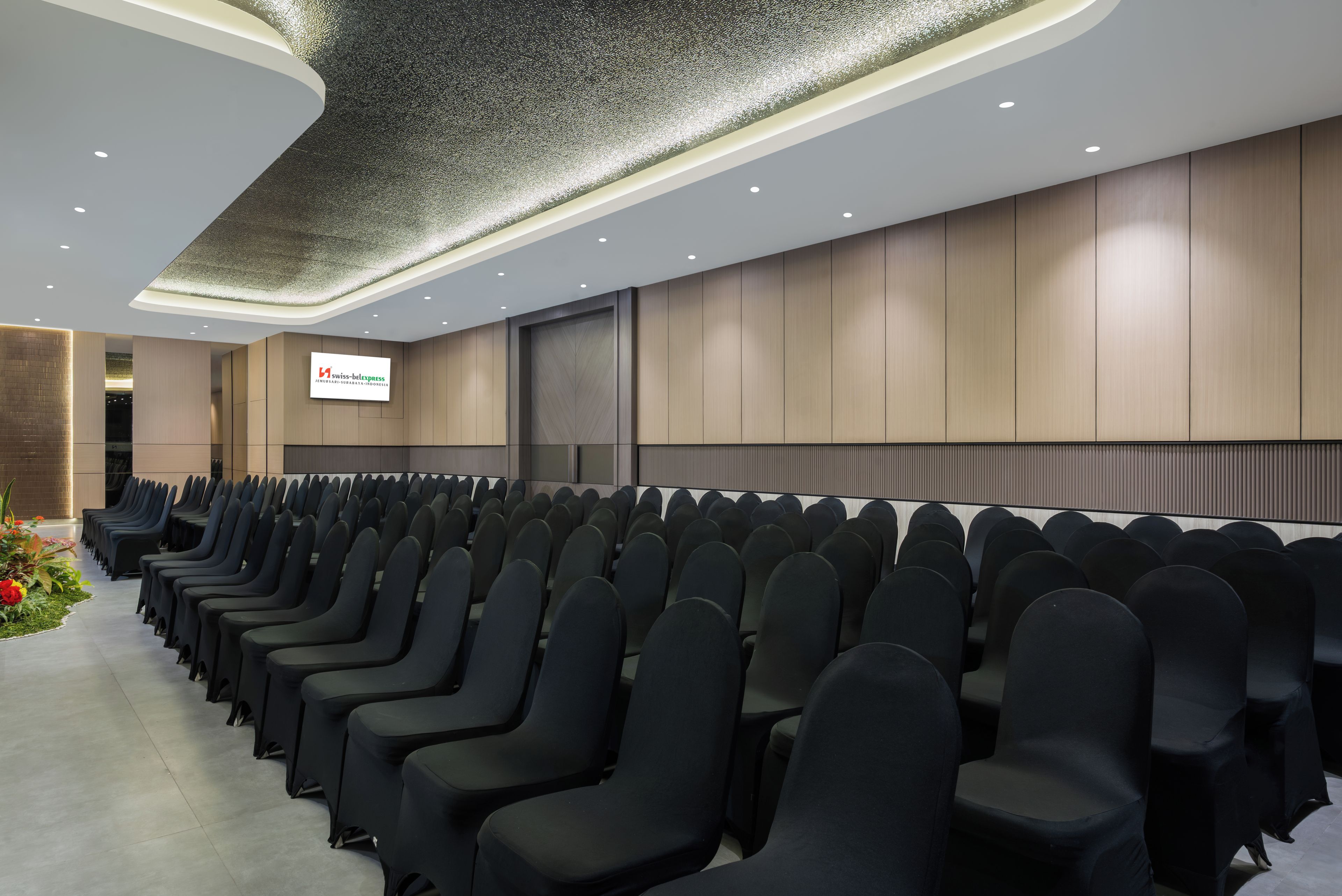Sala de conferência com fileiras de cadeiras, uma tela grande exibindo um logotipo e um design moderno no teto.