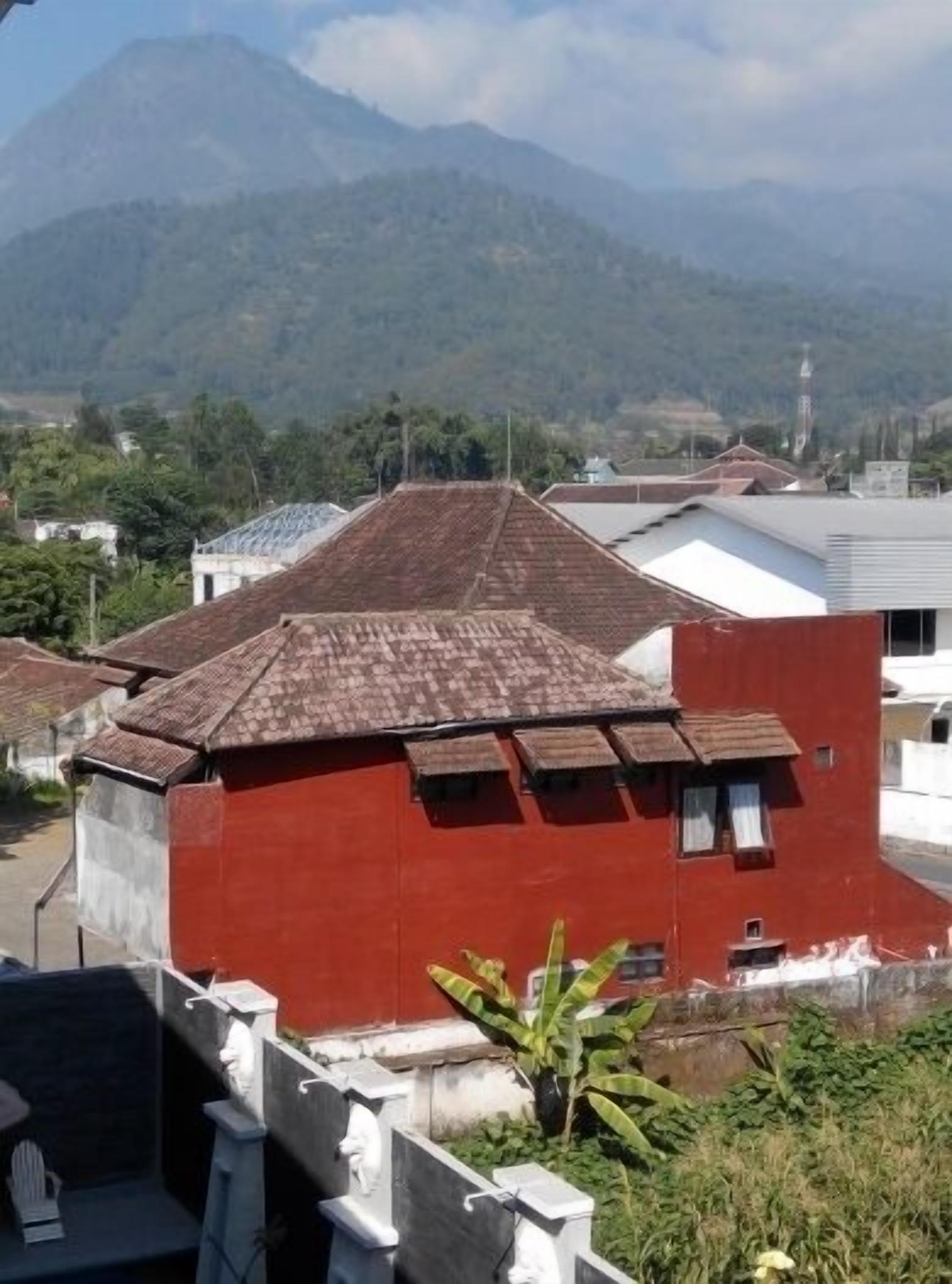Um edifício vermelho com telhado inclinado, rodeado por vegetação e uma montanha ao fundo.