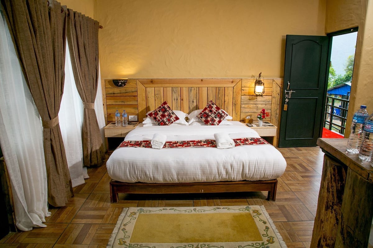 Quarto de hotel com cama de madeira, mesa de cabeceira com luminária, cabeceira de madeira, porta verde e piso de madeira.