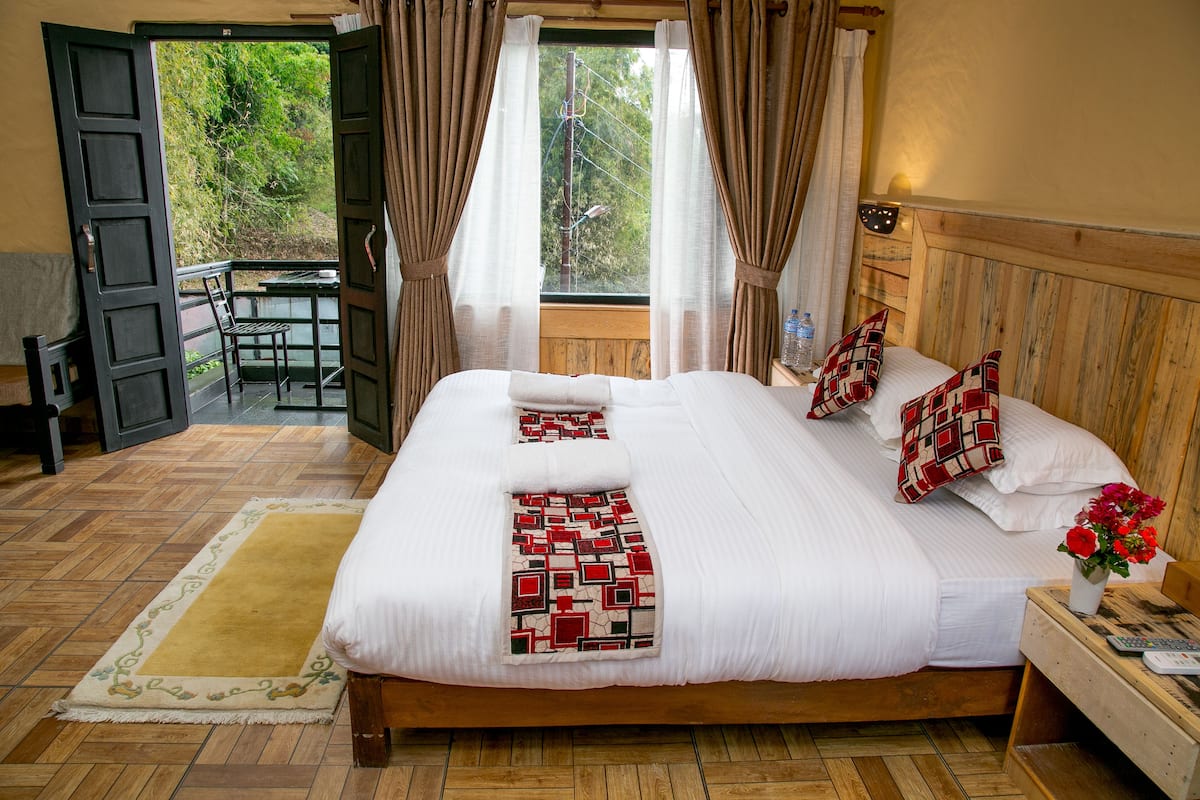 Quarto com cama de madeira, roupa de cama branca, uma mesinha com um controle remoto e uma janela com cortinas.