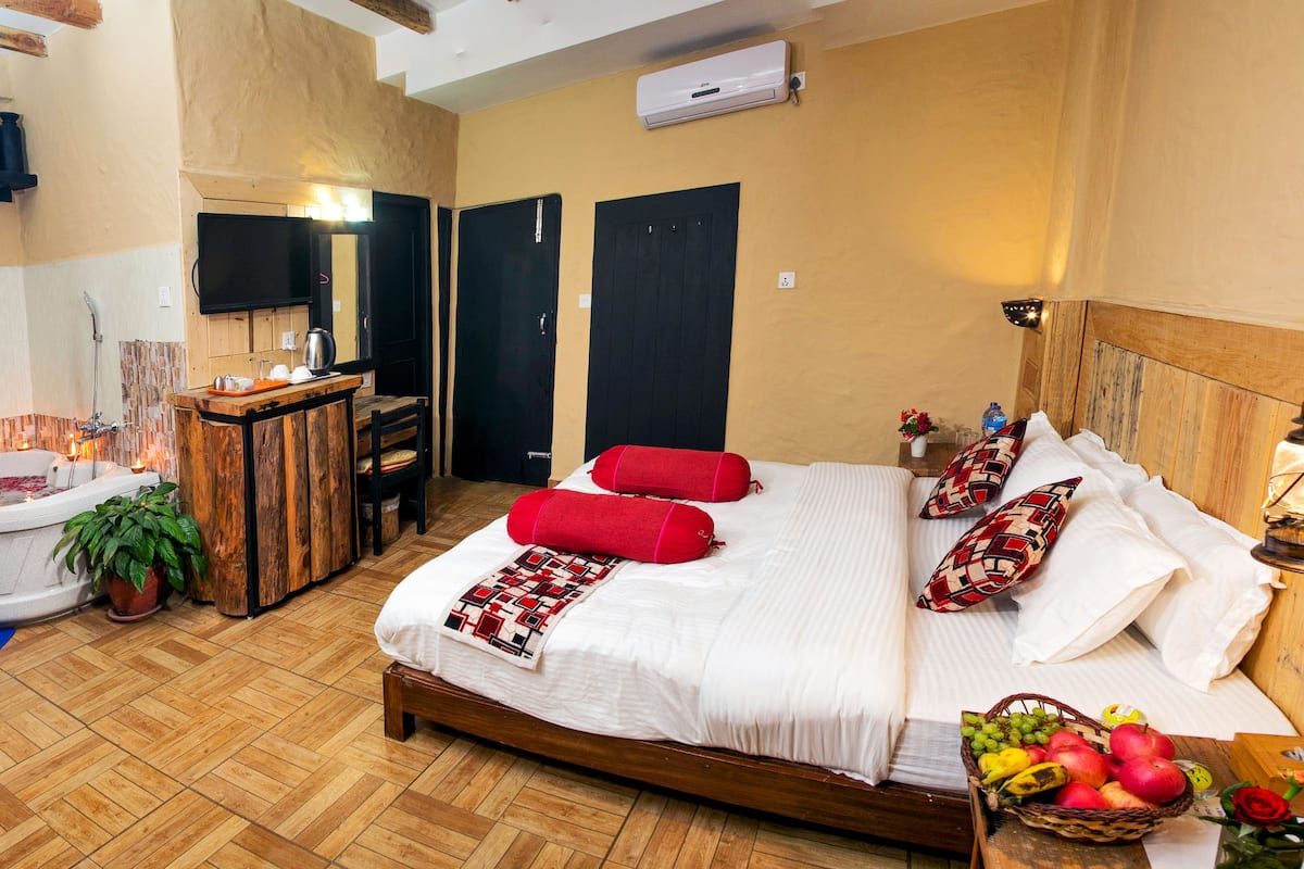 Quarto de hotel com piso de madeira, cama com lençóis brancos e almofadas vermelhas, mesa de cabeceira com abajur, cesta de frutas e banheiro com banheira e planta em vaso.