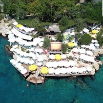 Vista aérea de um resort costeiro com vários níveis de áreas de estar ao ar livre, um edifício central e um píer que se estende para a água.