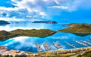 Uma marina com barcos atracados, cercada por colinas verdes exuberantes e um mar azul calmo sob um céu parcialmente nublado.