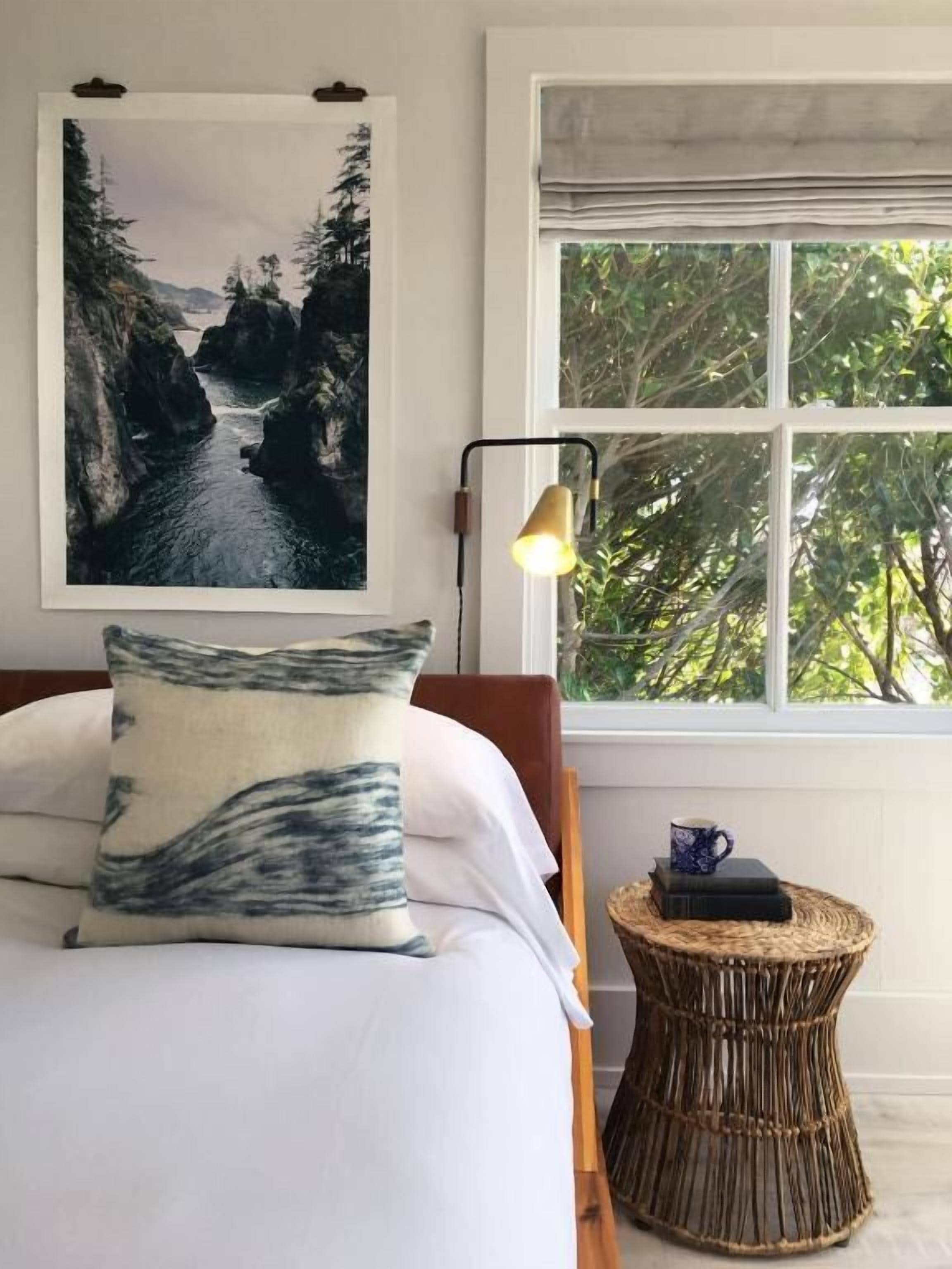 Quarto com cama, pintura pendurada na parede, abajur, mesinha de vime com uma caneca e janela com vista para árvores.