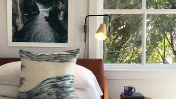 Quarto com cama, pintura pendurada na parede, abajur, mesinha de vime com uma caneca e janela com vista para árvores.