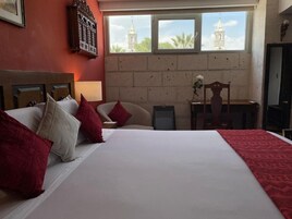 Quarto de hotel com uma cama grande, duas mesas de cabeceira, uma cadeira, uma escrivaninha e janela com vista para uma igreja.