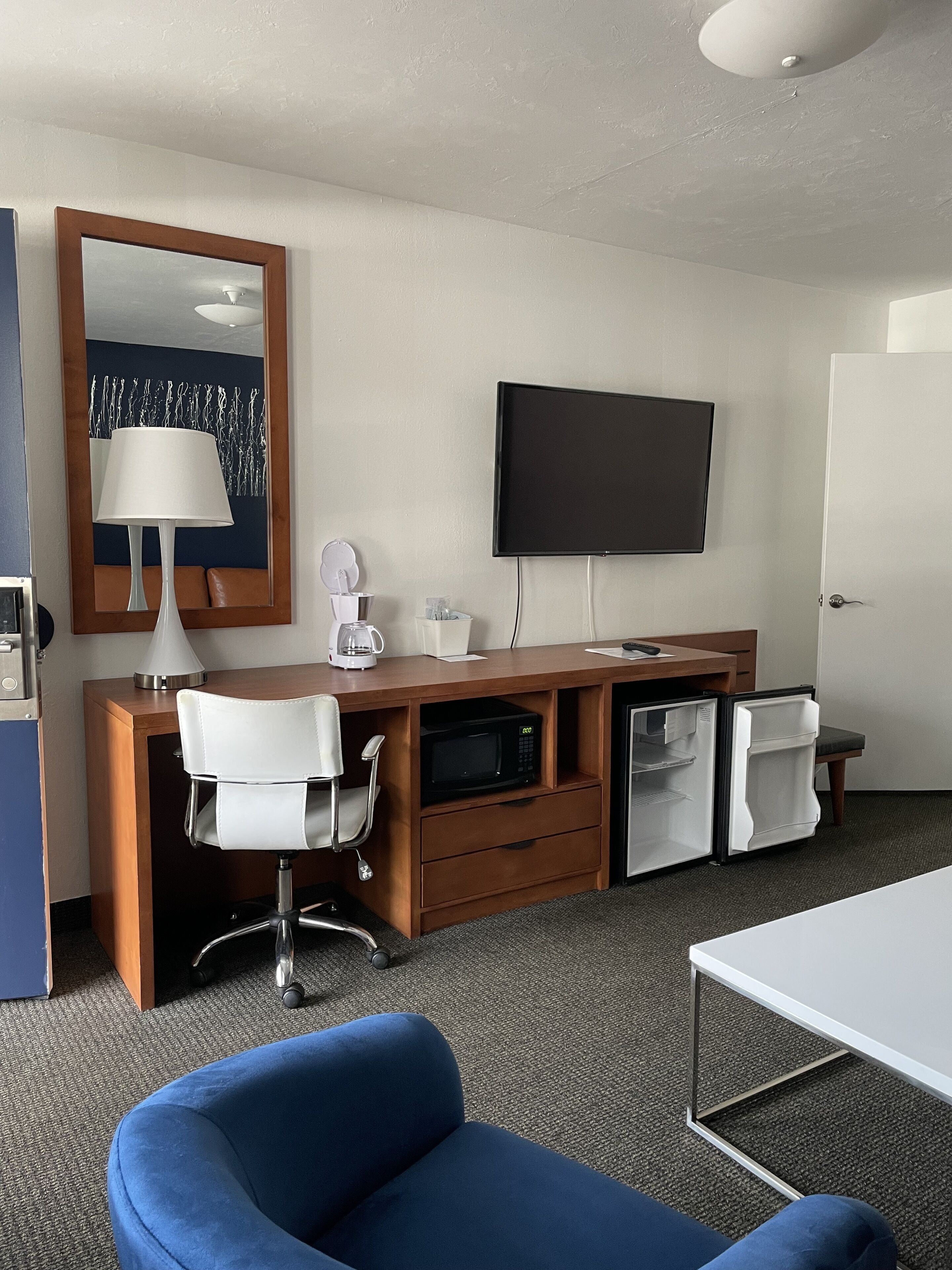 Quarto de hotel com escrivaninha, uma cadeira, televisão na parede, micro-ondas, frigorífico, máquina de café e espelho iluminado.