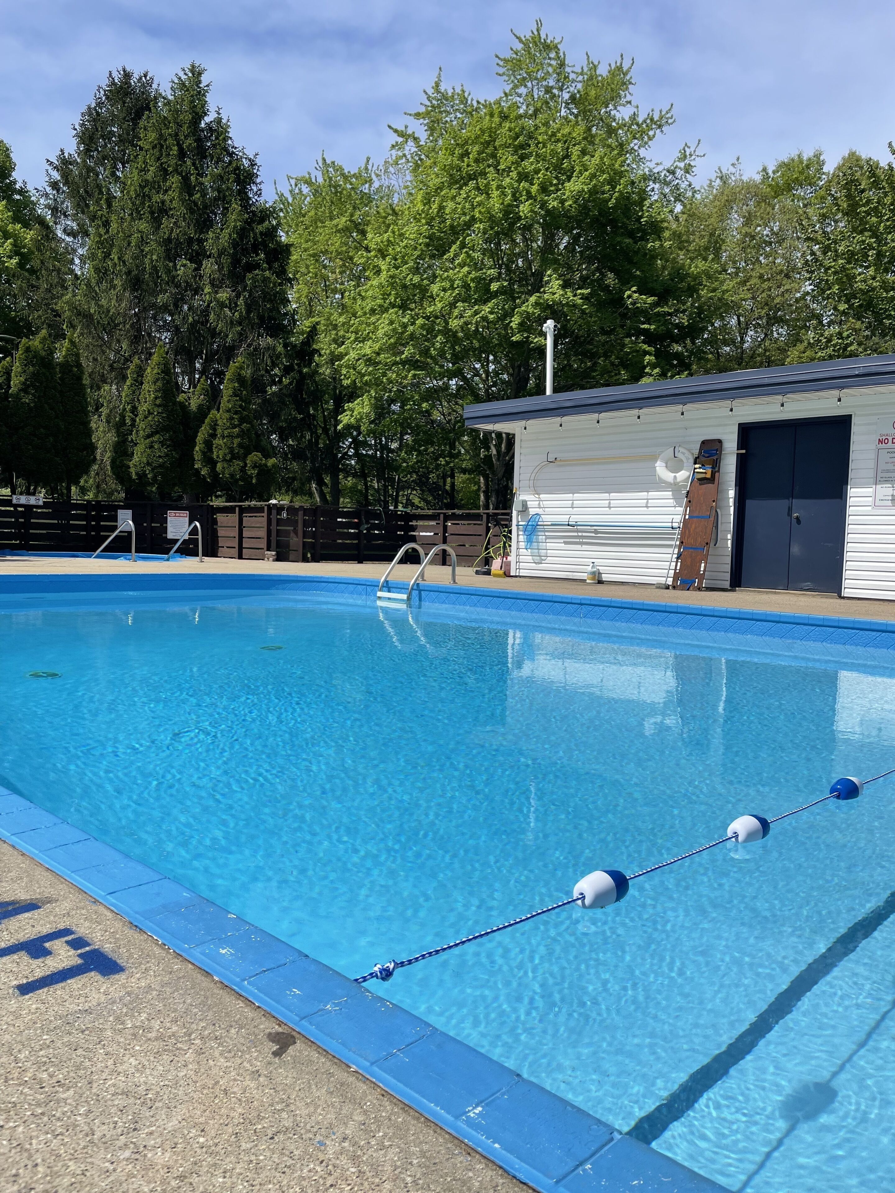 Piscina azul límpida com um deck de madeira, uma torre de salva-vidas e uma área cercada com árvores ao fundo.