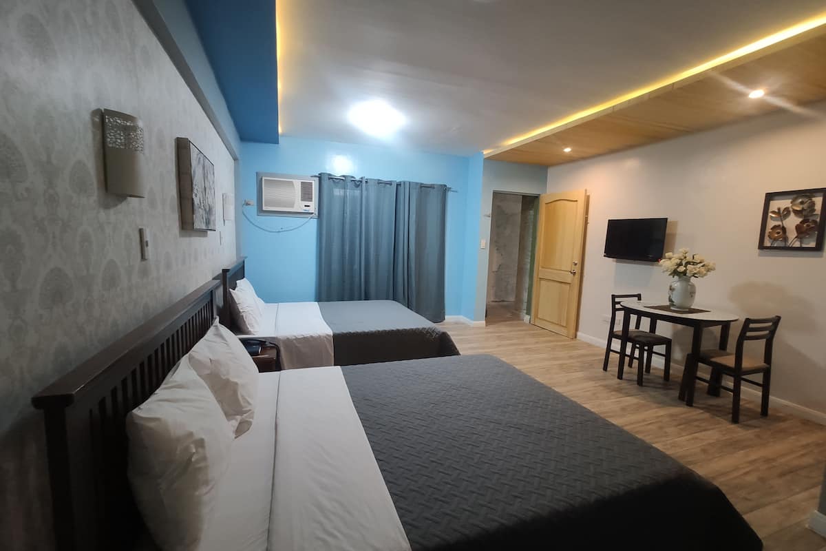 Quarto de hotel com duas camas, uma mesa com cadeiras, uma televisão e uma porta que dá para outro quarto.