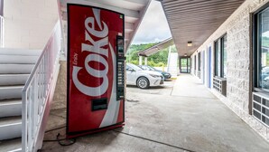Máquina de Coca-Cola fora de um edifício com galeria coberta e carros estacionados.