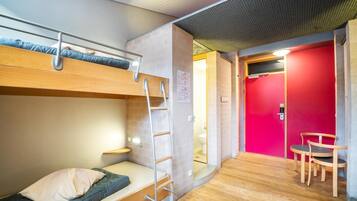 Quarto compacto com beliches, uma mesinha e parede vermelha.