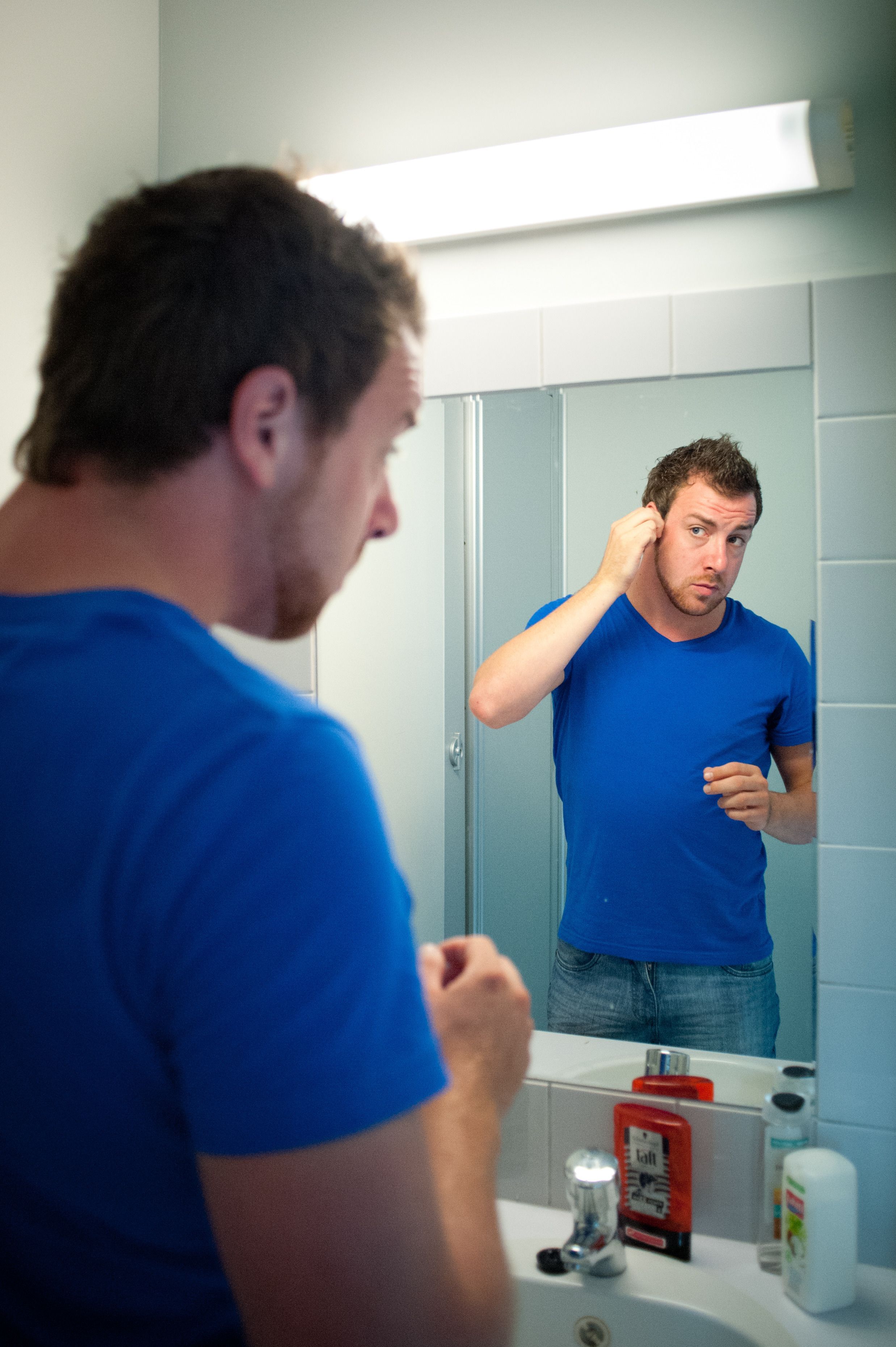 Um homem de camisa azul em frente a um espelho de banheiro, escovando o cabelo.