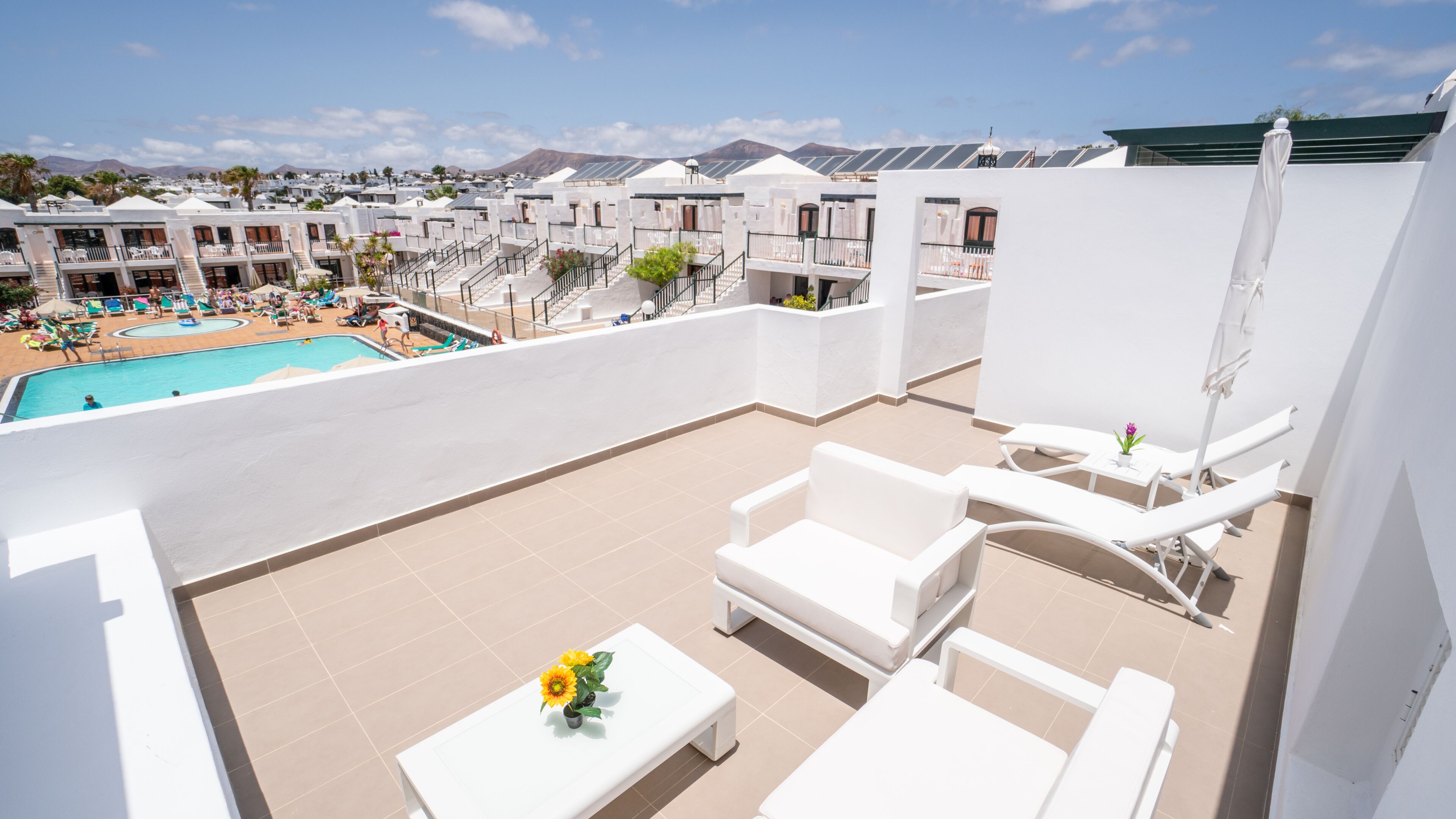 Uma área de piscina no terraço com cadeiras de lounge brancas, uma pequena mesa com uma flor e vista para um resort com vários edifícios e uma montanha ao fundo.