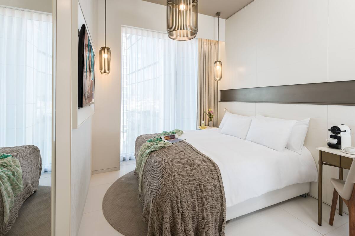 Quarto de hotel moderno com uma cama grande, uma mesinha de cabeceira com cafeteira e vista para a cidade através de amplas janelas.