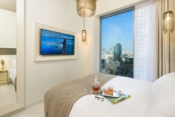 Quarto de hotel moderno com uma janela grande que oferece vista para a cidade, uma cama bem arrumada, uma mesa de cabeceira com uma garrafa e copos, e um quadro pendurado na parede.