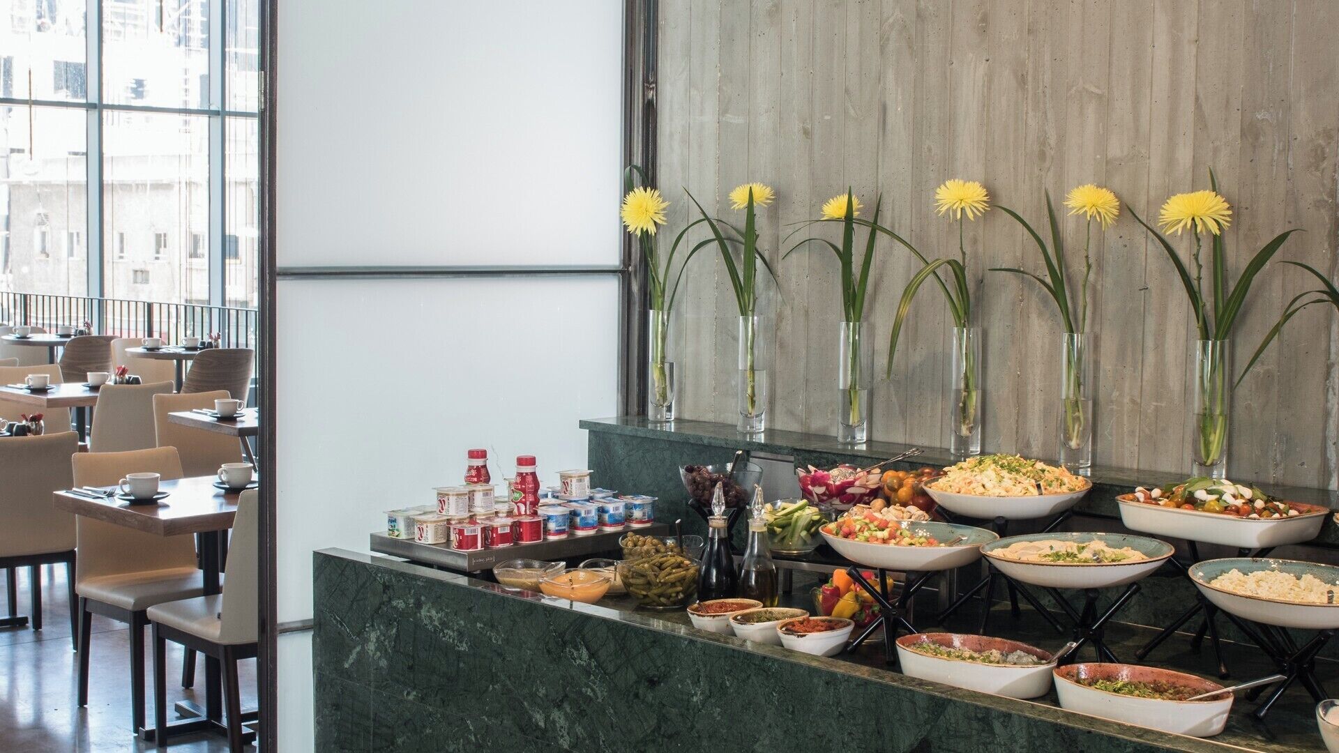Disposição de buffet com diversos alimentos e acompanhamentos, área de refeições com cadeiras e uma ampla janela com vista para a paisagem urbana.