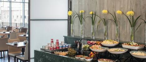 Disposição de buffet com diversos alimentos e acompanhamentos, área de refeições com cadeiras e uma ampla janela com vista para a paisagem urbana.