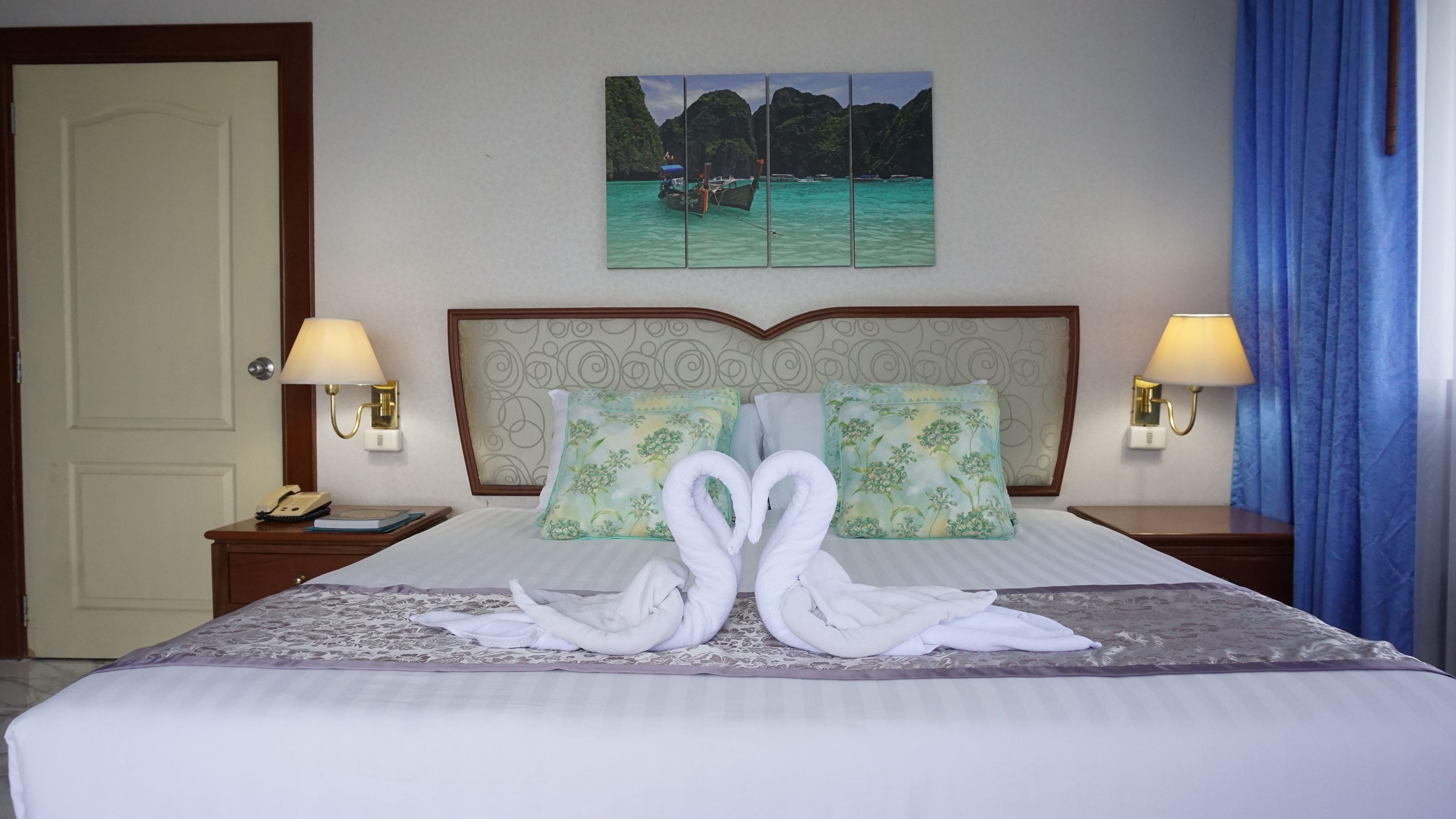 Quarto de hotel com cama, exibe toalha disposta em formato de coração, almofadas estampadas com flores e pintura de um barco na água acima da cama.