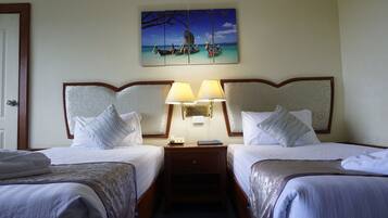 Quarto de hotel com duas camas, uma mesa de cabeceira com abajur, um quadro de barcos numa praia e uma porta.