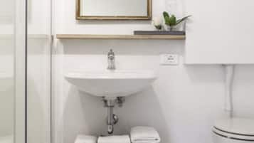 Casa de banho com lavatório branco, espelho com moldura dourada, pequena prateleira com uma planta e um porta-toalhas.