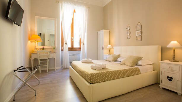 Quarto com cama, mesa de cabeceira, luminária, escrivaninha, cadeira e televisão fixada na parede.