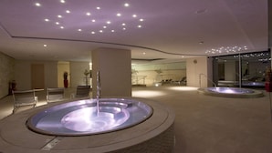 Uma área interna de piscina moderna, com uma banheira de hidromassagem central, assentos ao redor da piscina e uma parede de vidro ao fundo.