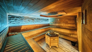 Uma sauna com bancos de madeira e piso ladrilhado, uma pequena mesa de madeira com uma tigela, e um painel de vidro com vista para o exterior.