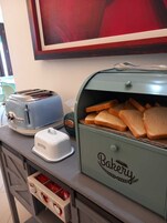 Uma bancada de cozinha com uma torradeira, um recipiente para manteiga e uma caixa de pão com a etiqueta 'Padaria'.