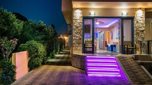 Entrada de hotel moderna com porta de vidro, escadaria com iluminação roxa e um jardim bem cuidado.