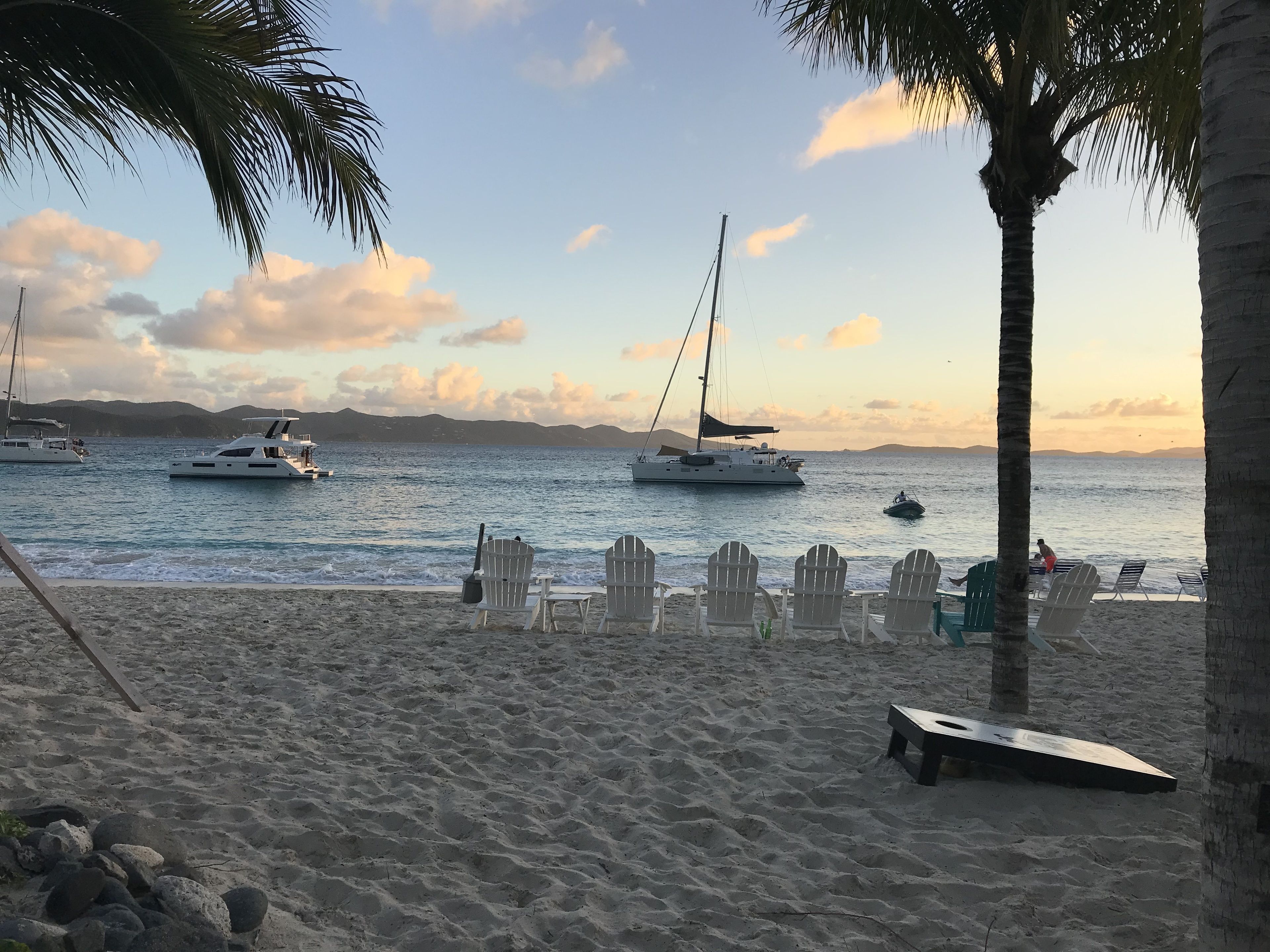 Uma praia com cadeiras brancas e um banco, um veleiro e um jet ski na água.