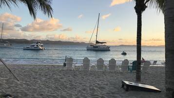 Uma praia com cadeiras brancas e um banco, um veleiro e um jet ski na água.