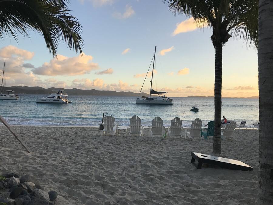 Uma praia com cadeiras brancas e um banco, um veleiro e um jet ski na água.