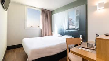 Quarto de hotel com uma cama, uma escrivaninha com um laptop, uma cadeira e uma janela com cortinas.