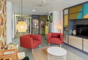 Um saguão moderno com poltronas vermelhas, uma mesa de centro branca e um balcão de recepção com um quiosque digital.