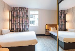 Quarto de hotel com duas camas, um espelho grande e uma janela com cortinas.