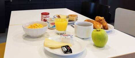 Uma mesa de café da manhã com uma tigela de cereal, um copo de suco de laranja, um croissant, uma maçã verde e um prato com uma fatia de queijo e uma barra de chocolate pequena.