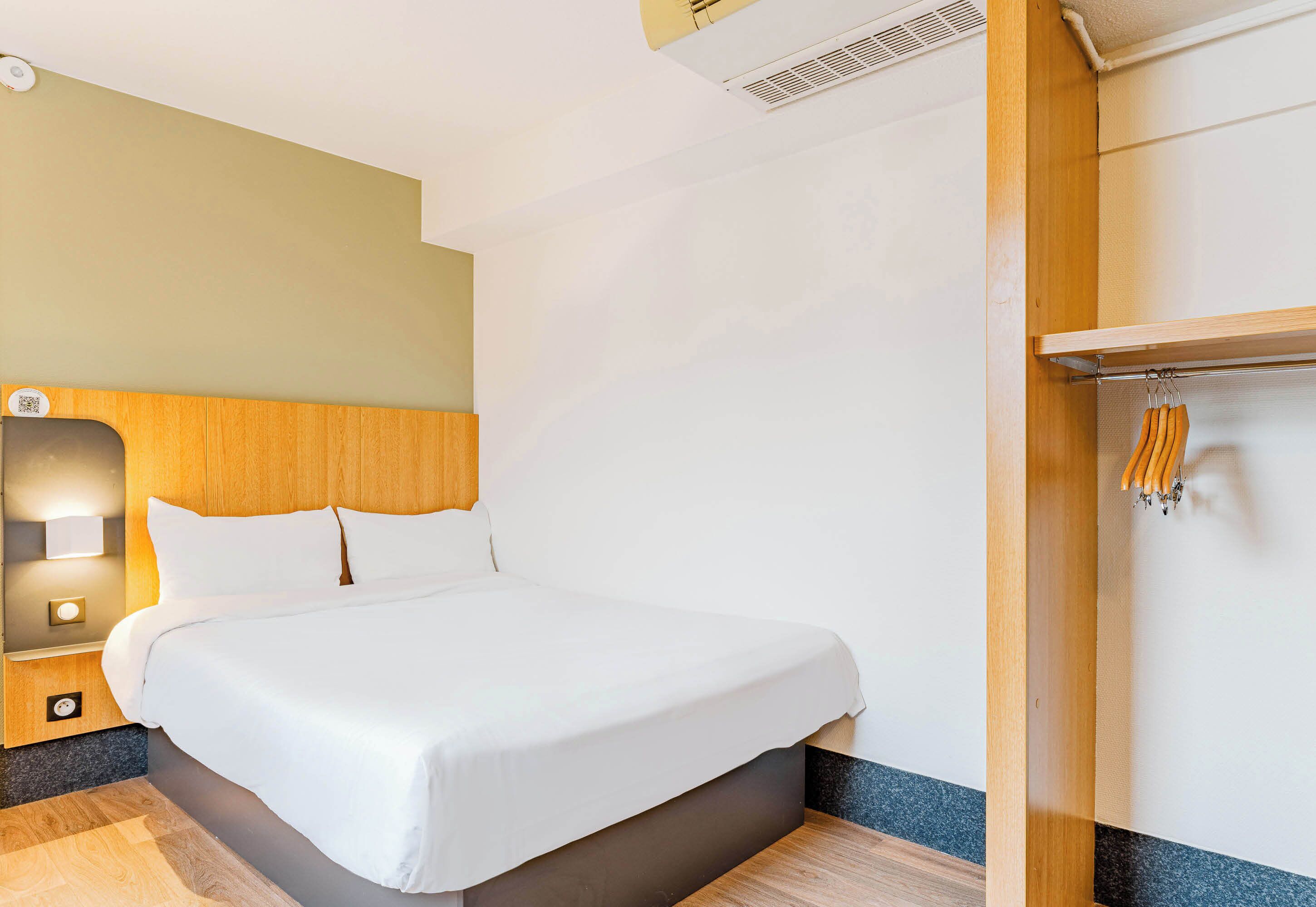 Quarto de hotel com uma cama, cabeceira de madeira, mesa de cabeceira, luminária na parede e um cabideiro com ganchos.